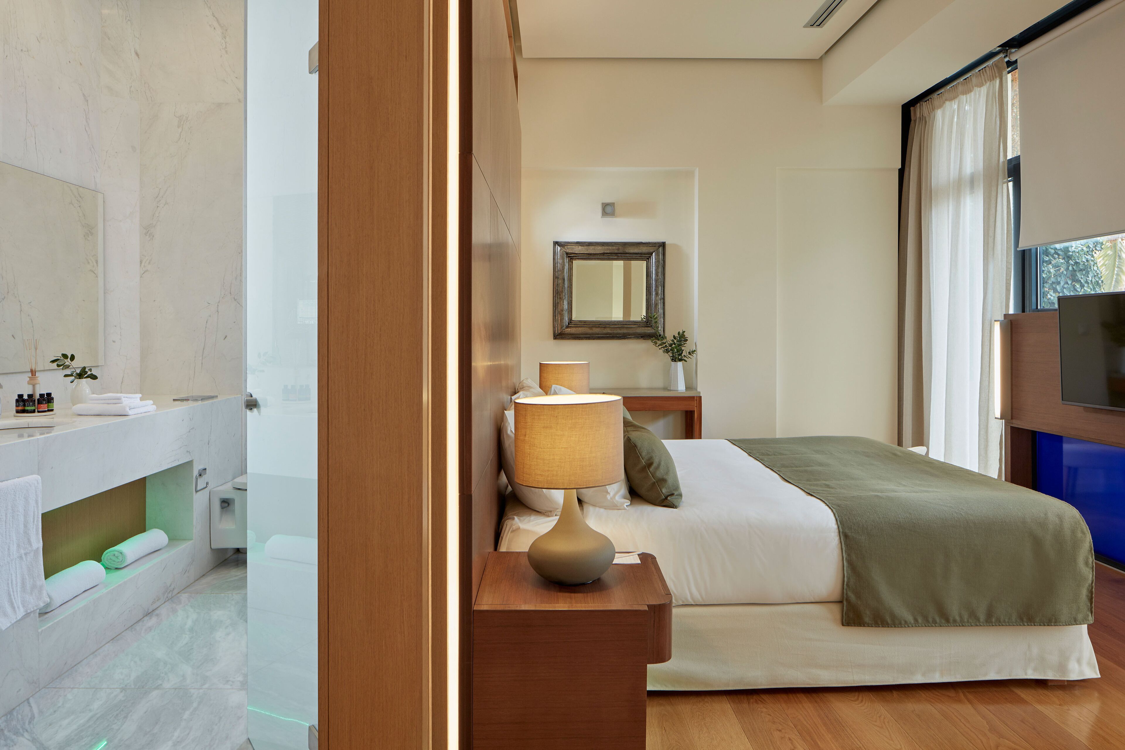Quarto de hotel moderno com casa de banho em mármore, uma luminária de cabeceira, cama com manta verde e televisão na parede direita.