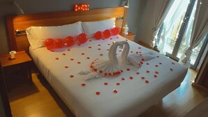 Cama bem arrumada com toalha em formato de cisne, pétalas de rosa vermelhas, balões vermelhos e mesa de cabeceira com abajur.