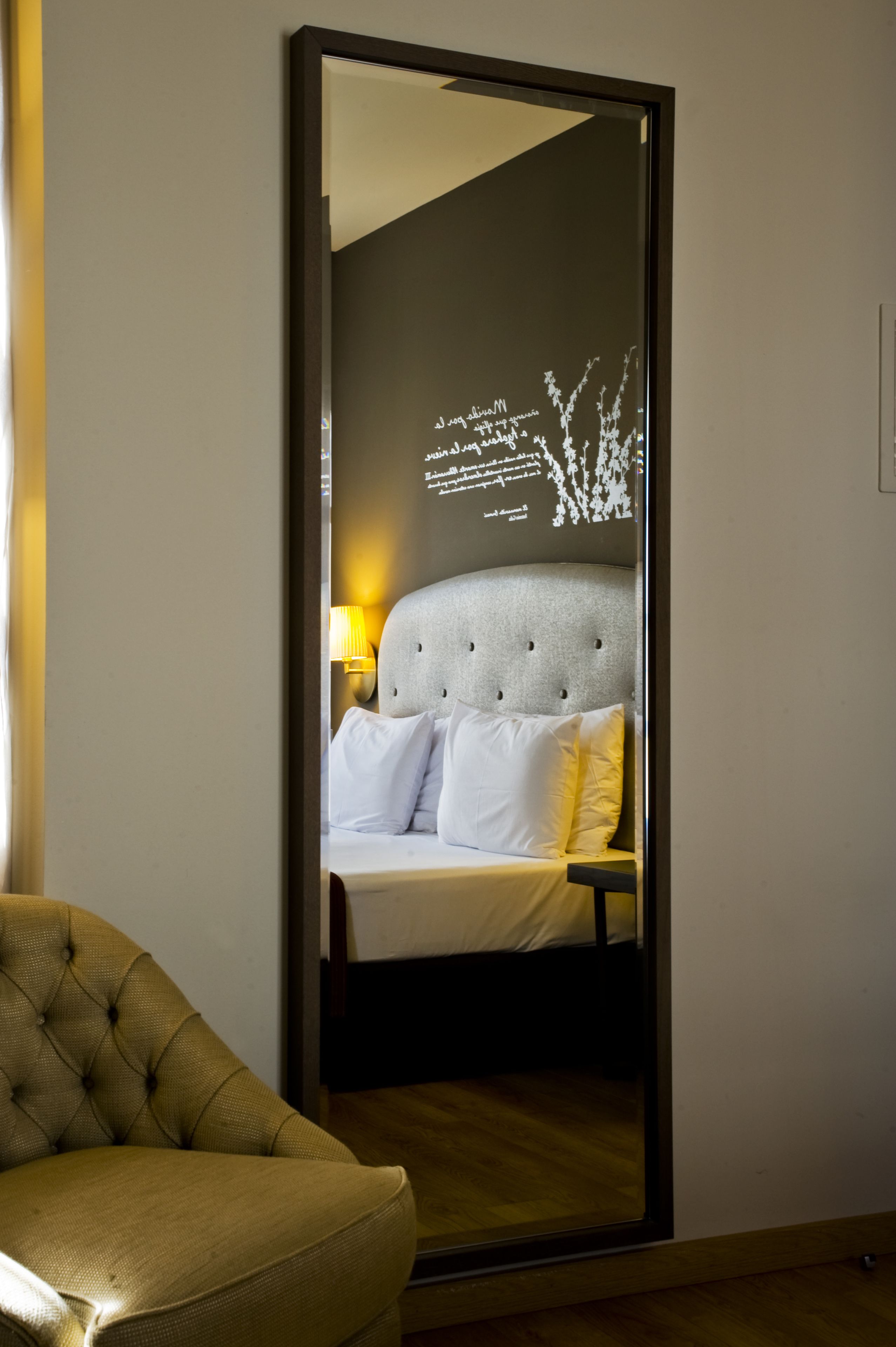 Quarto com cama, cadeira e espelho fixado na parede refletindo o ambiente.