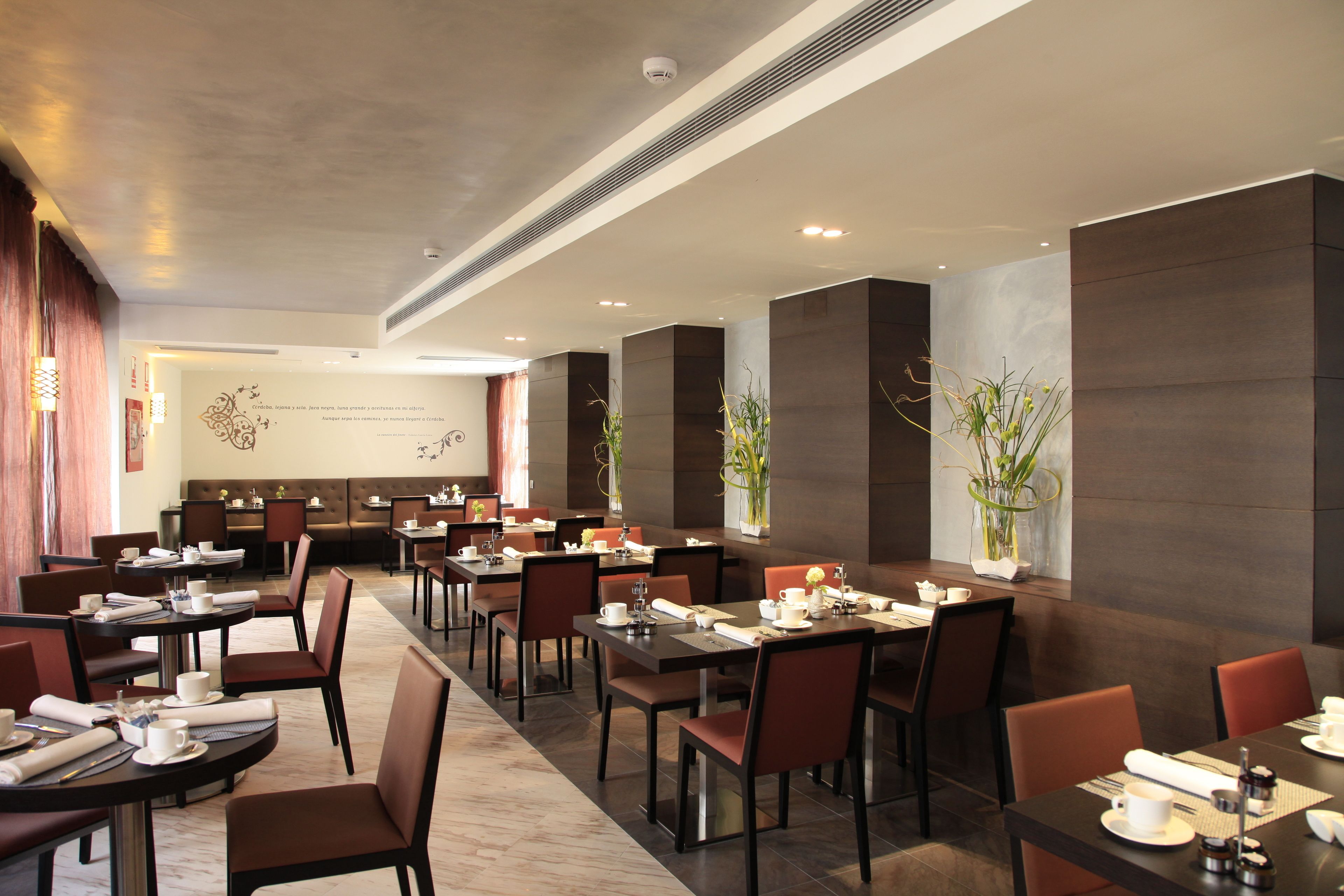 Uma sala de jantar moderna com várias mesas e cadeiras, um espaço de estar central e plantas decorativas.