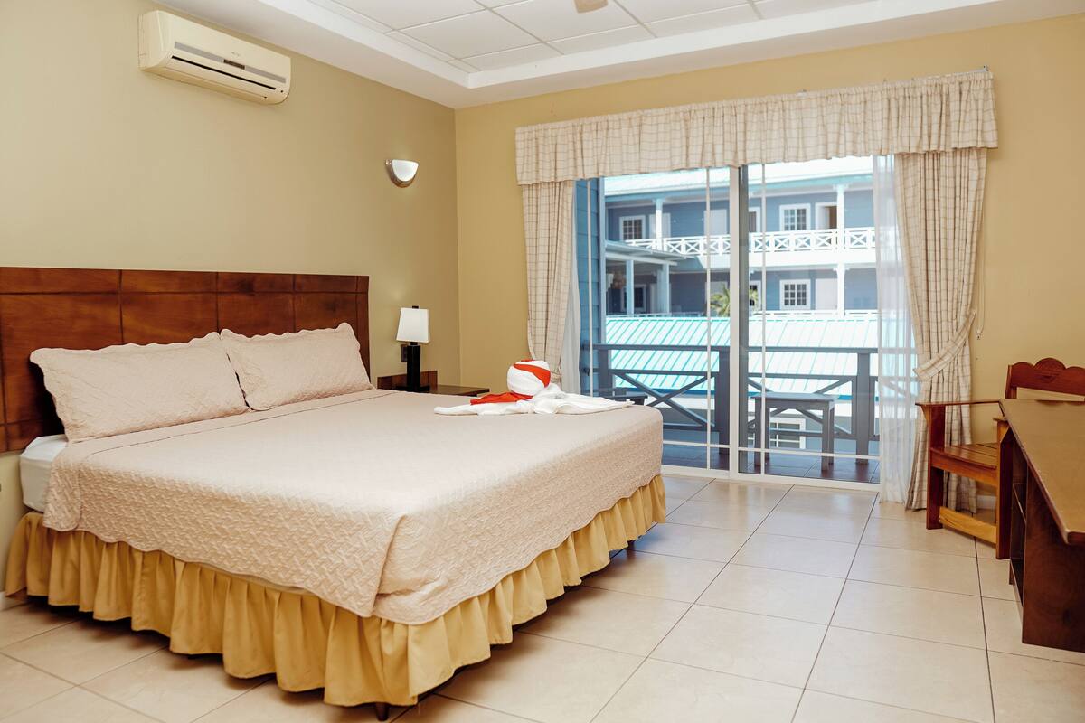 Quarto de hotel com uma cama grande, mesa de cabeceira, luminária, varanda com vista para edifícios e ar condicionado.