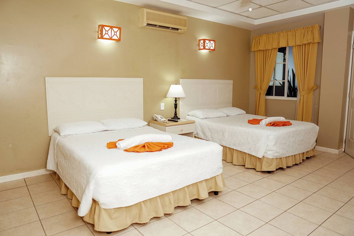Quarto de hotel com duas camas, uma mesa de cabeceira, uma luminária e uma janela com cortinas.
