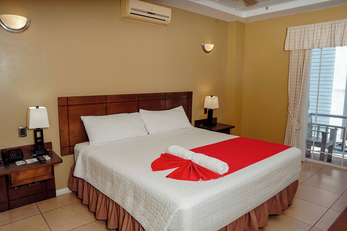 Uma cama bem arrumada com uma toalha vermelha e um travesseiro branco, uma mesa de cabeceira de madeira com uma luminária e uma janela com cortinas.