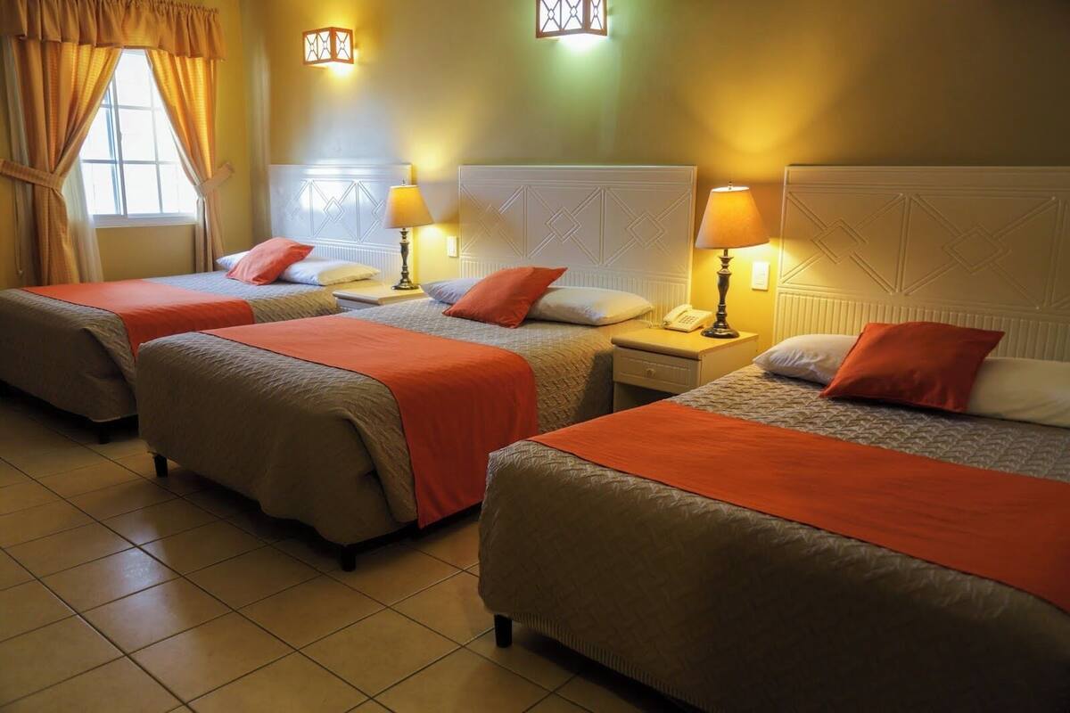 Quarto de hotel com três camas de solteiro, cada uma com cobertor e travesseiro vermelhos. Há luminárias e telefone nas mesas de cabeceira.