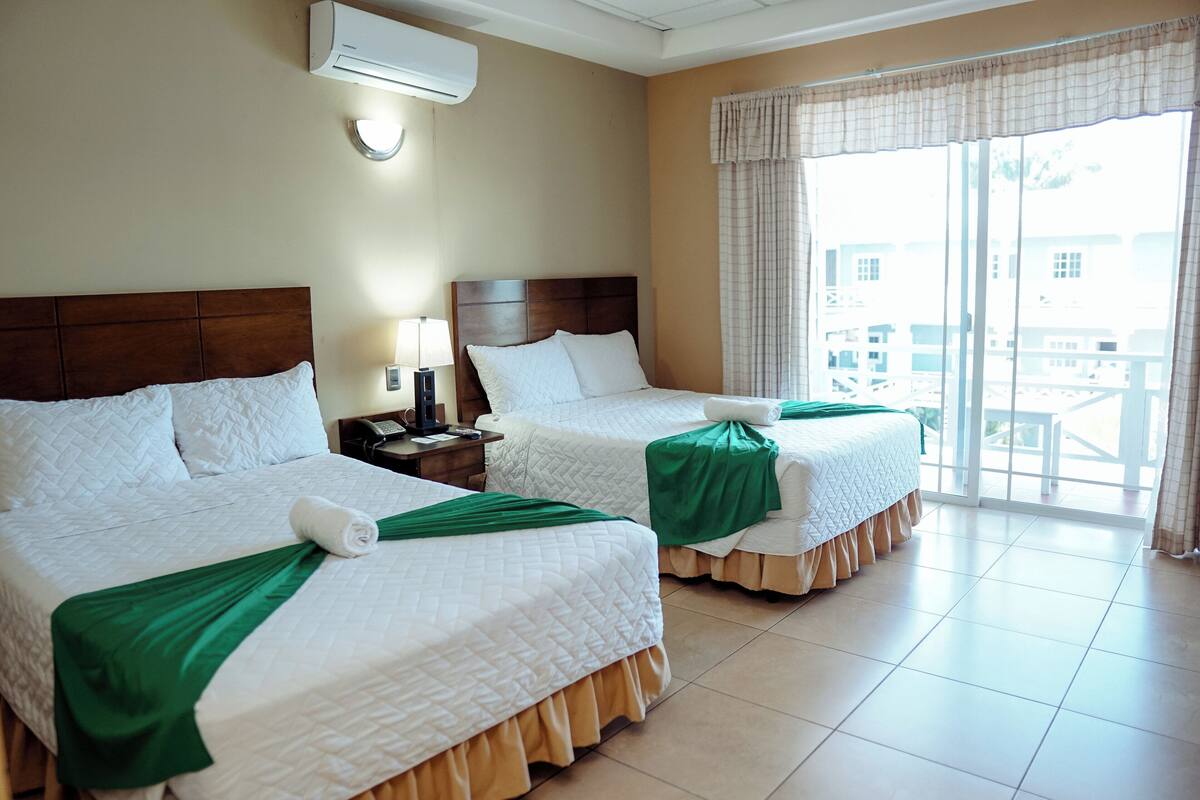 Quarto de hotel com duas camas, uma mesinha de cabeceira, uma luminária e vista para o exterior.