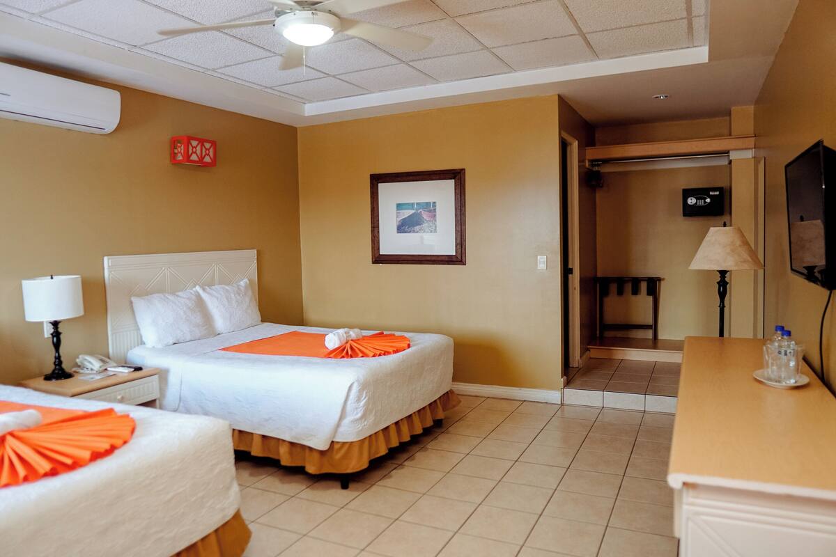 Quarto de hotel com duas camas, um ventilador de teto, ar condicionado, um quadro pendurado na parede, mesa de cabeceira com abajur e escrivaninha com uma garrafa de água.