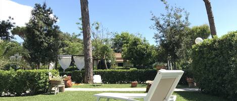 Um jardim com duas cadeiras de descanso brancas, um gramado bem cuidado e diversas árvores e arbustos.