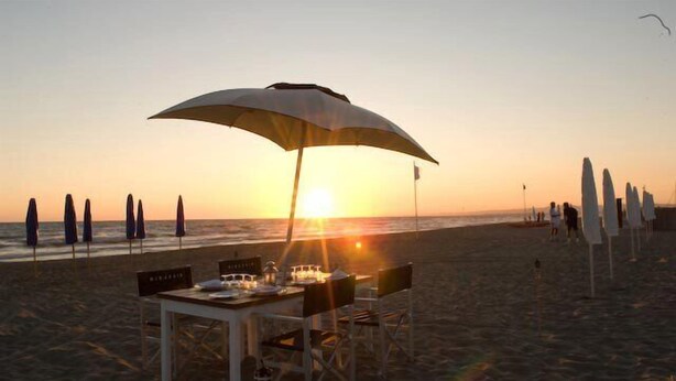 Disposição de refeições à beira-mar, com mesa e cadeiras sob um guarda-sol, com vista para o oceano ao pôr do sol.