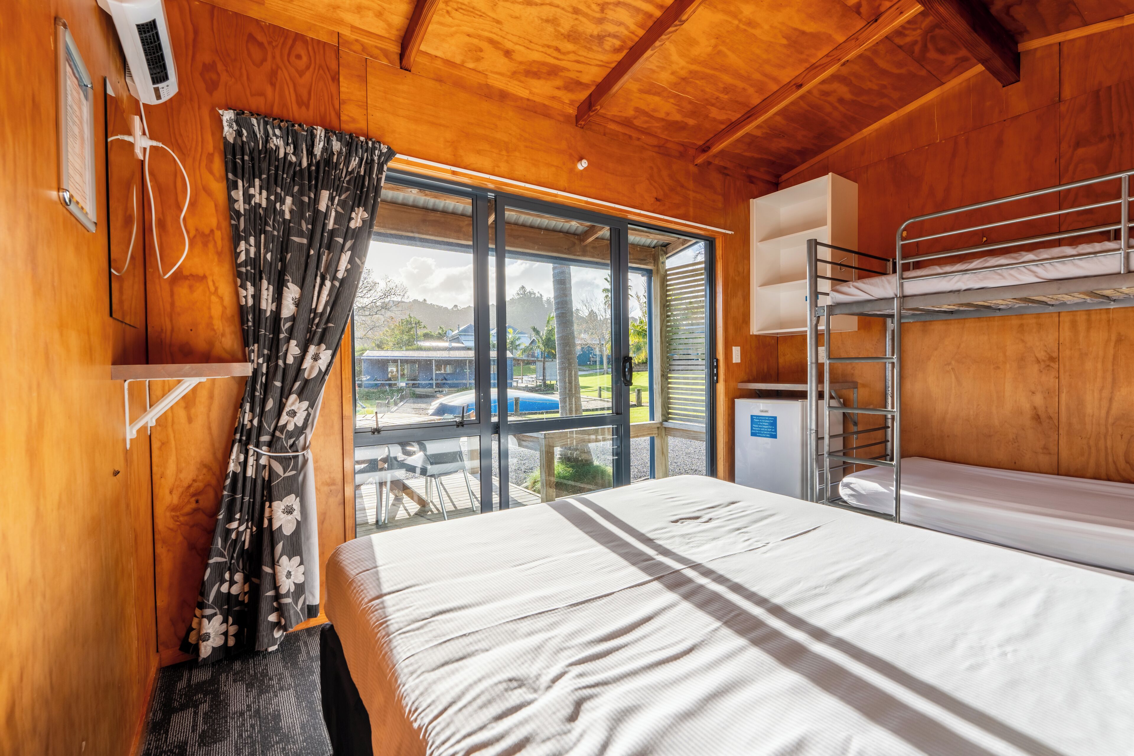 Quarto de cabana de madeira com beliche, escrivaninha com cadeira, janela com cortinas e vista para o exterior.