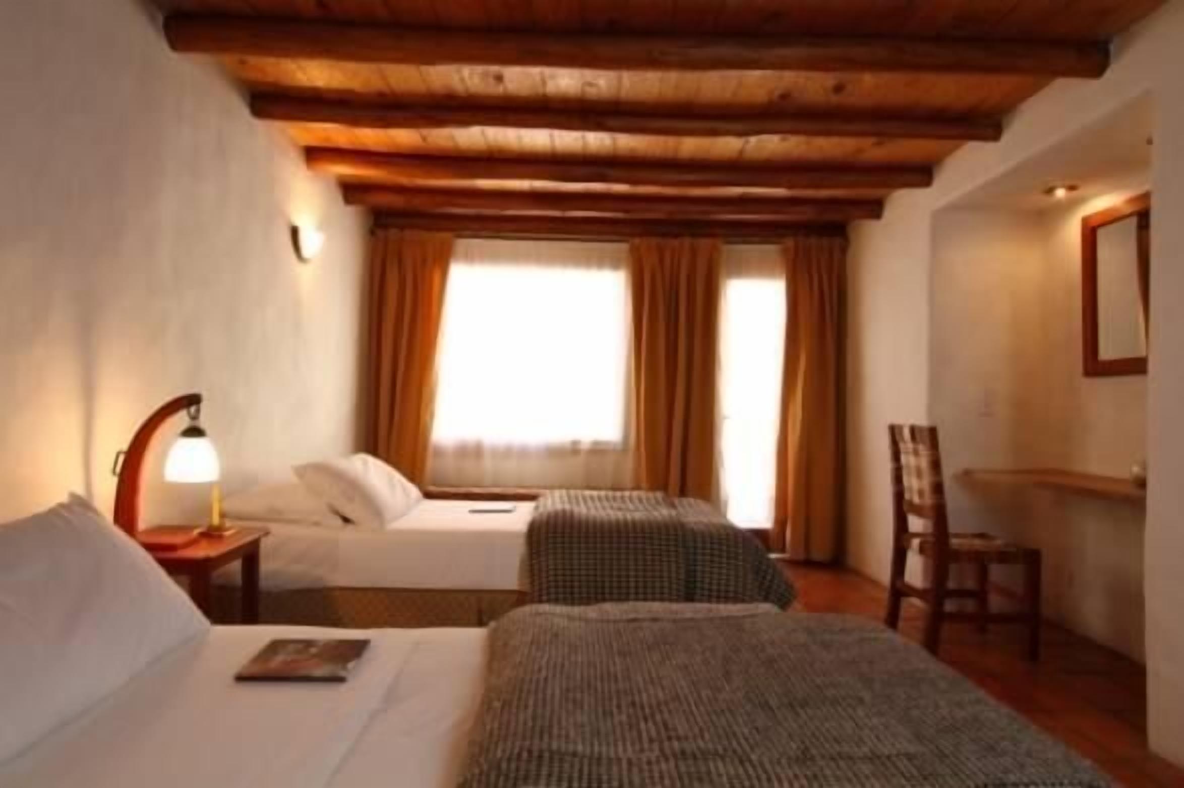 Quarto de hotel com duas camas, uma escrivaninha, uma cadeira e uma janela com cortinas.