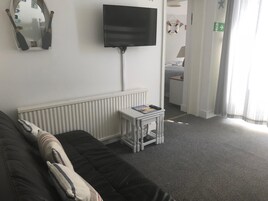 Uma sala de estar com um sofá preto, uma pequena mesa de apoio branca com um livro, uma TV de tela plana na parede e um radiador branco.