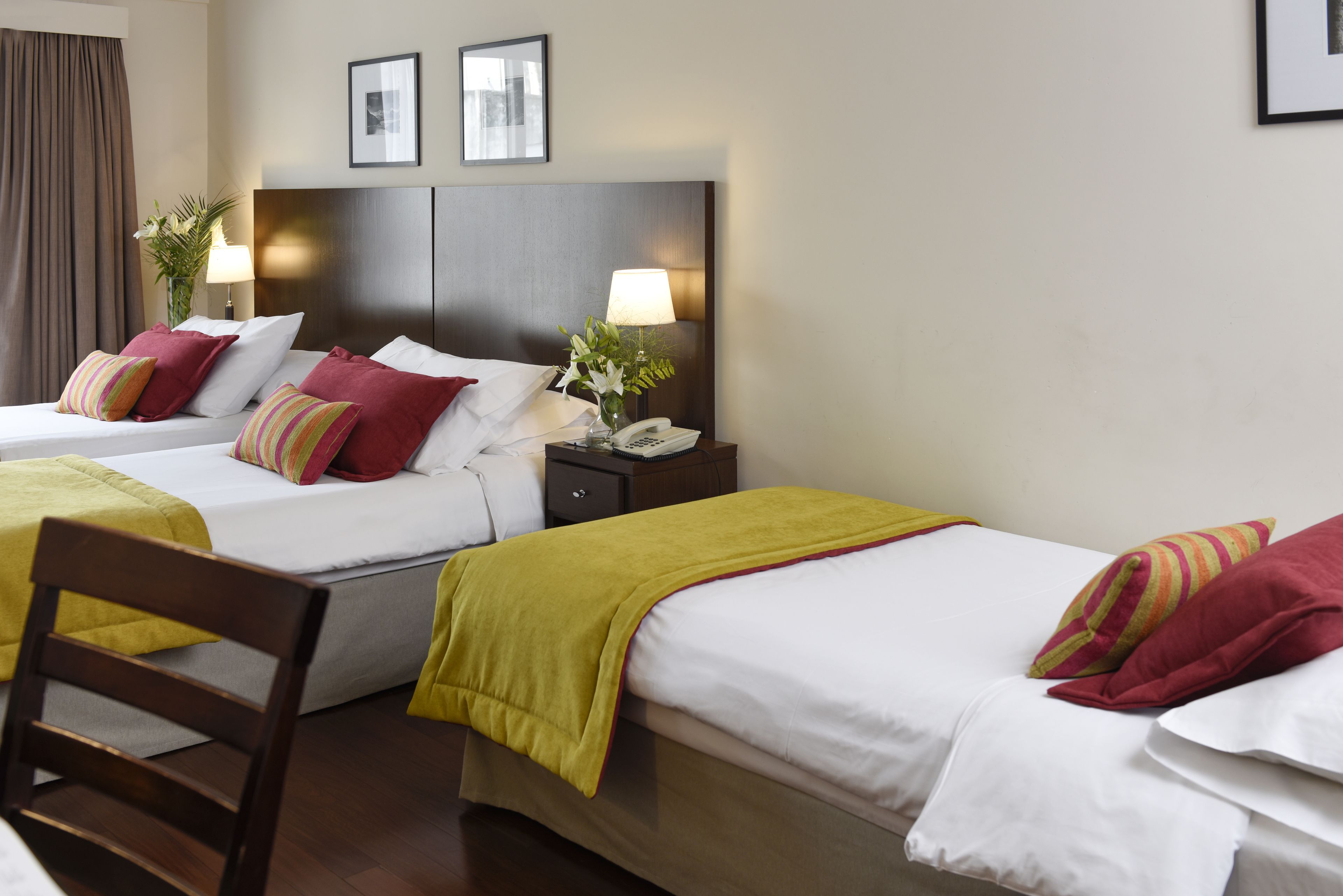 Quarto de hotel com duas camas, uma cadeira de madeira, uma mesa de cabeceira com abajur e um quadro na parede.