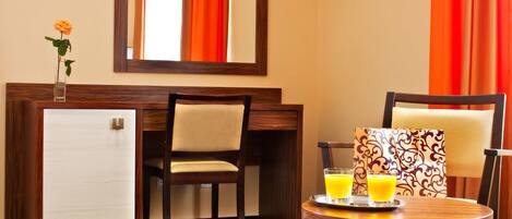 Quarto de hotel com escrivaninha de madeira, pequeno frigorífico, uma cadeira, mesa com bandeja de sumo de laranja e espelho na parede.