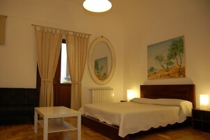 Quarto com cama, mesa de cabeceira, cadeira, janela com cortinas, radiador, pintura na parede e abajur no criado-mudo.
