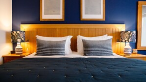 Cama bem arrumada com um edredom azul escuro, duas almofadas e uma cabeceira de madeira. Há duas obras de arte emolduradas na parede acima da cama.