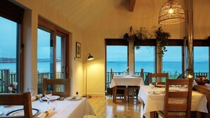 Uma área de refeições com mesas preparadas para dois, grandes janelas com vista para o mar e um lustre.