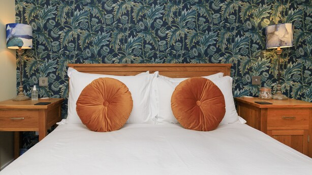 Uma cama bem arrumada com duas almofadas grandes alaranjadas, uma cabeceira de madeira e um padrão de papel de parede floral. Há duas mesas de cabeceira com abajures e um criado-mudo com uma luminária.