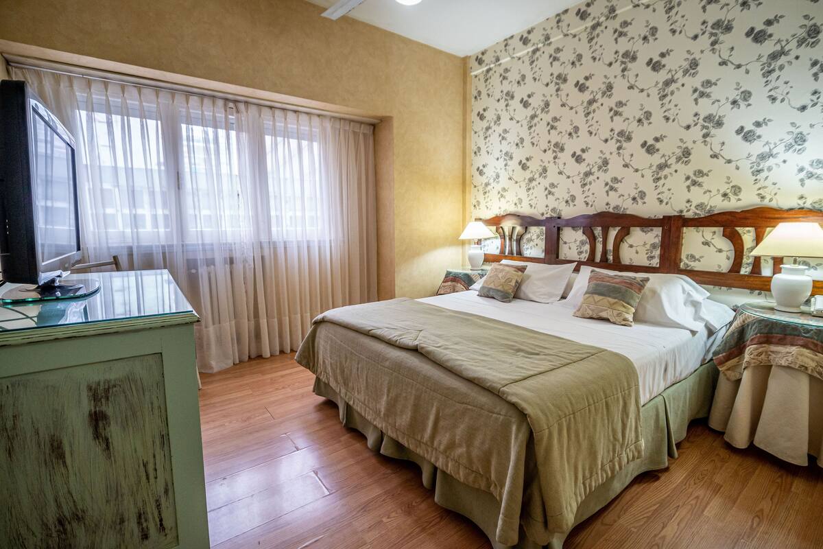 Quarto de hotel com cama, mesas de cabeceira, escrivaninha com computador e janela com cortinas.
