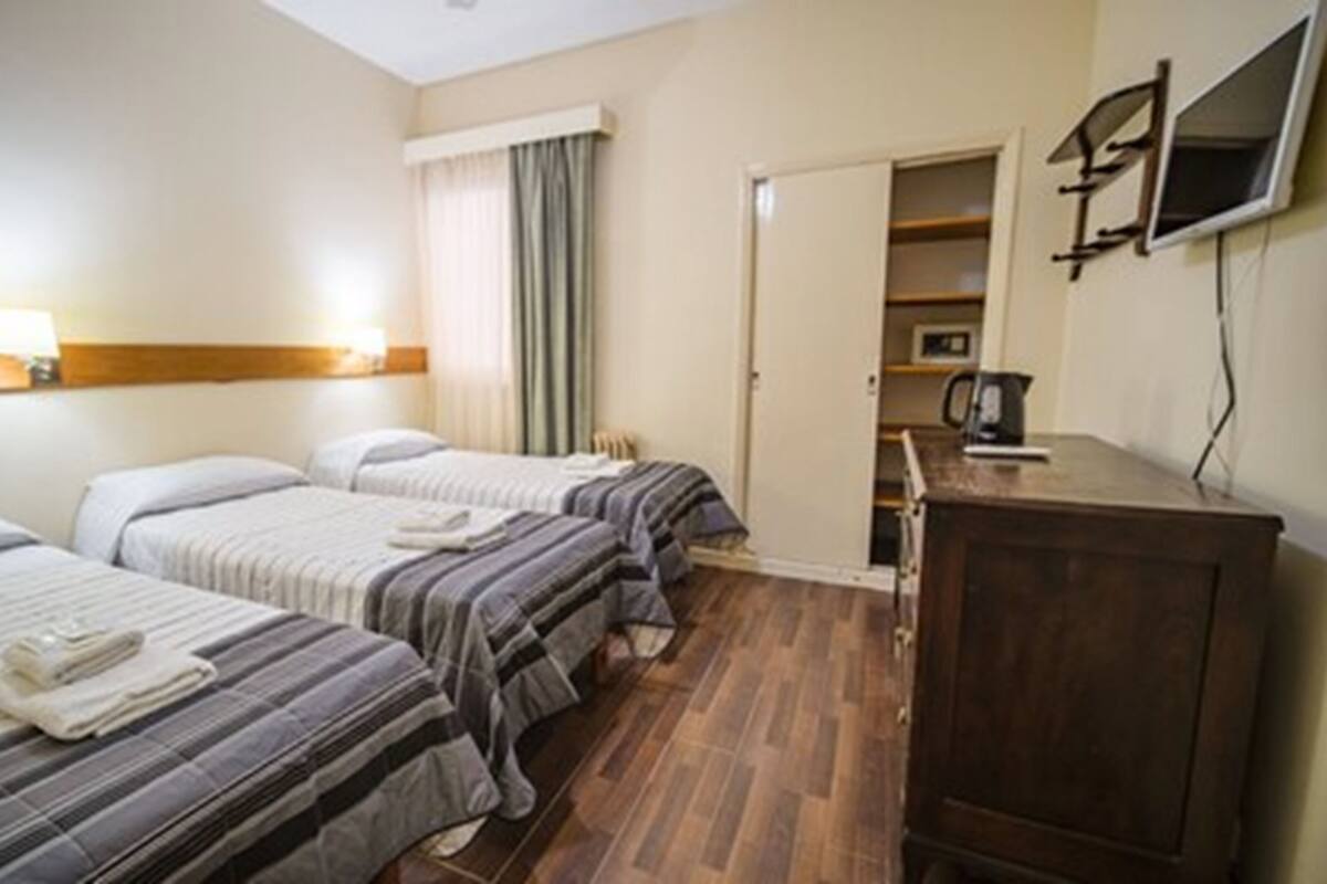 Quarto de hotel com três camas, cabeceira de madeira, televisão de tela plana na parede e um armário de madeira.