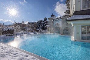 Uma piscina com água azul cristalina, rodeada por árvores cobertas de neve e um edifício com fachada de pedra.