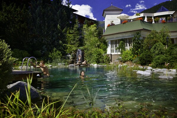 Uma piscina cristalina rodeada por vegetação exuberante, um deque de madeira e um edifício com uma placa que diz 'Wellness School'.