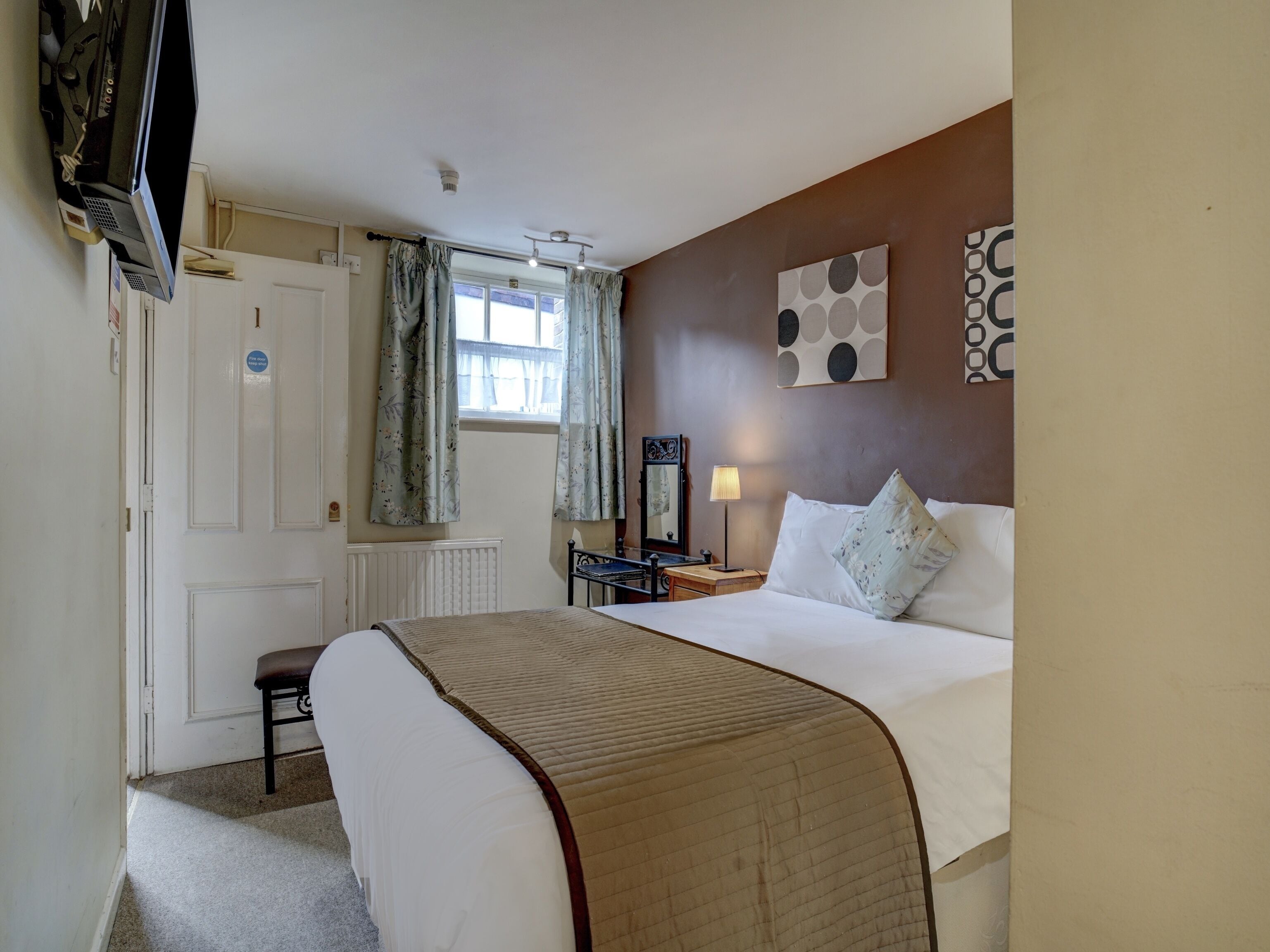 Quarto de hotel com cama, escrivaninha, cadeira, TV fixada na parede e janela com cortinas.