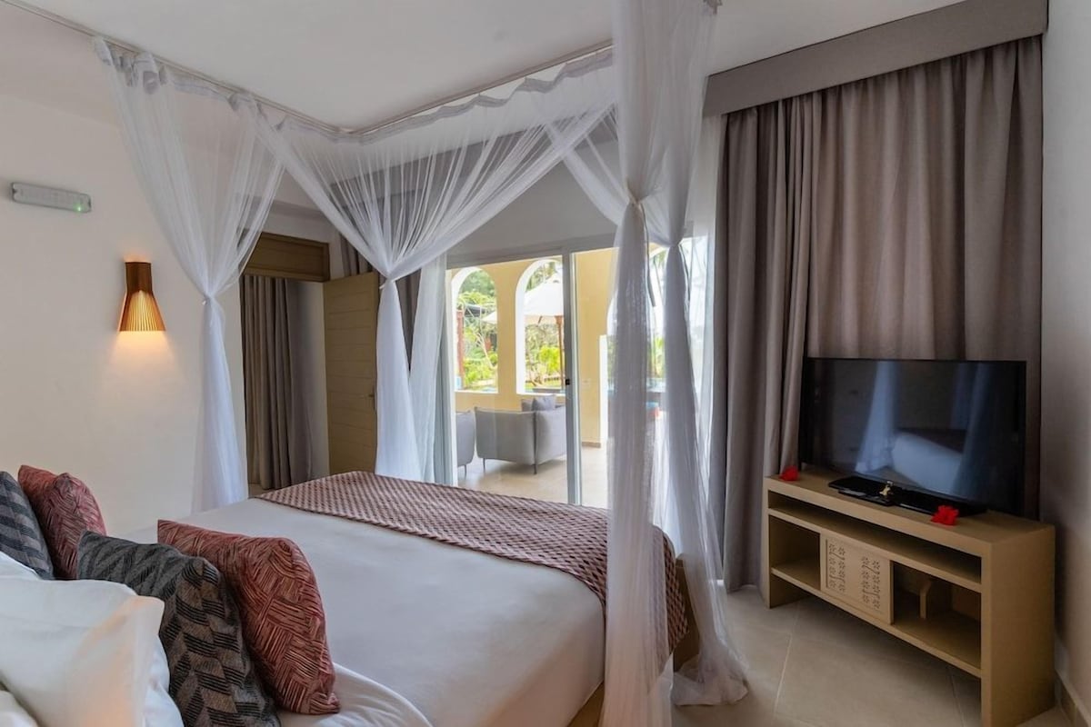 Quarto com cama, televisão sobre um suporte de madeira e vista para o exterior.