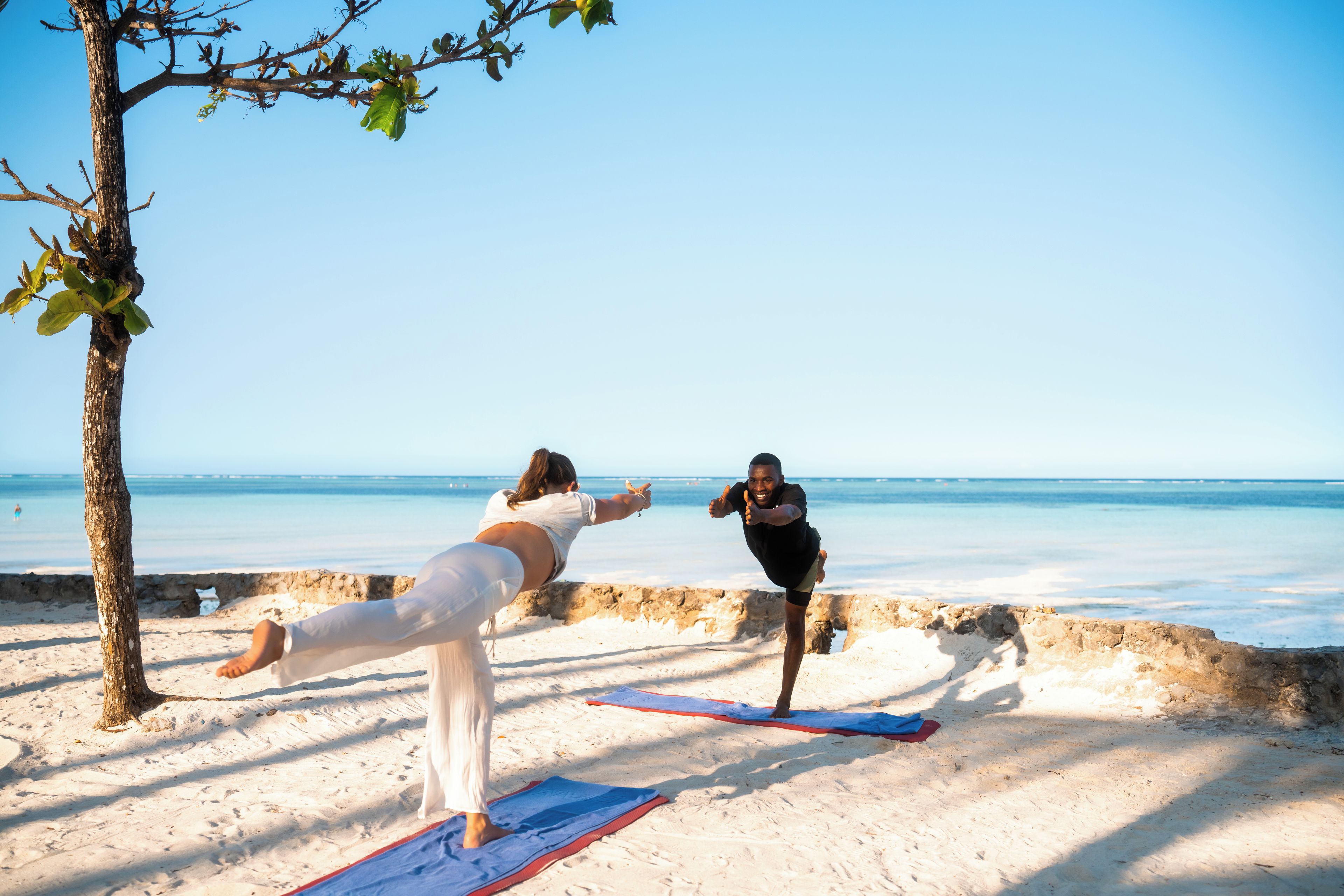 Duas pessoas praticando yoga numa praia, com um céu claro e oceano ao fundo.