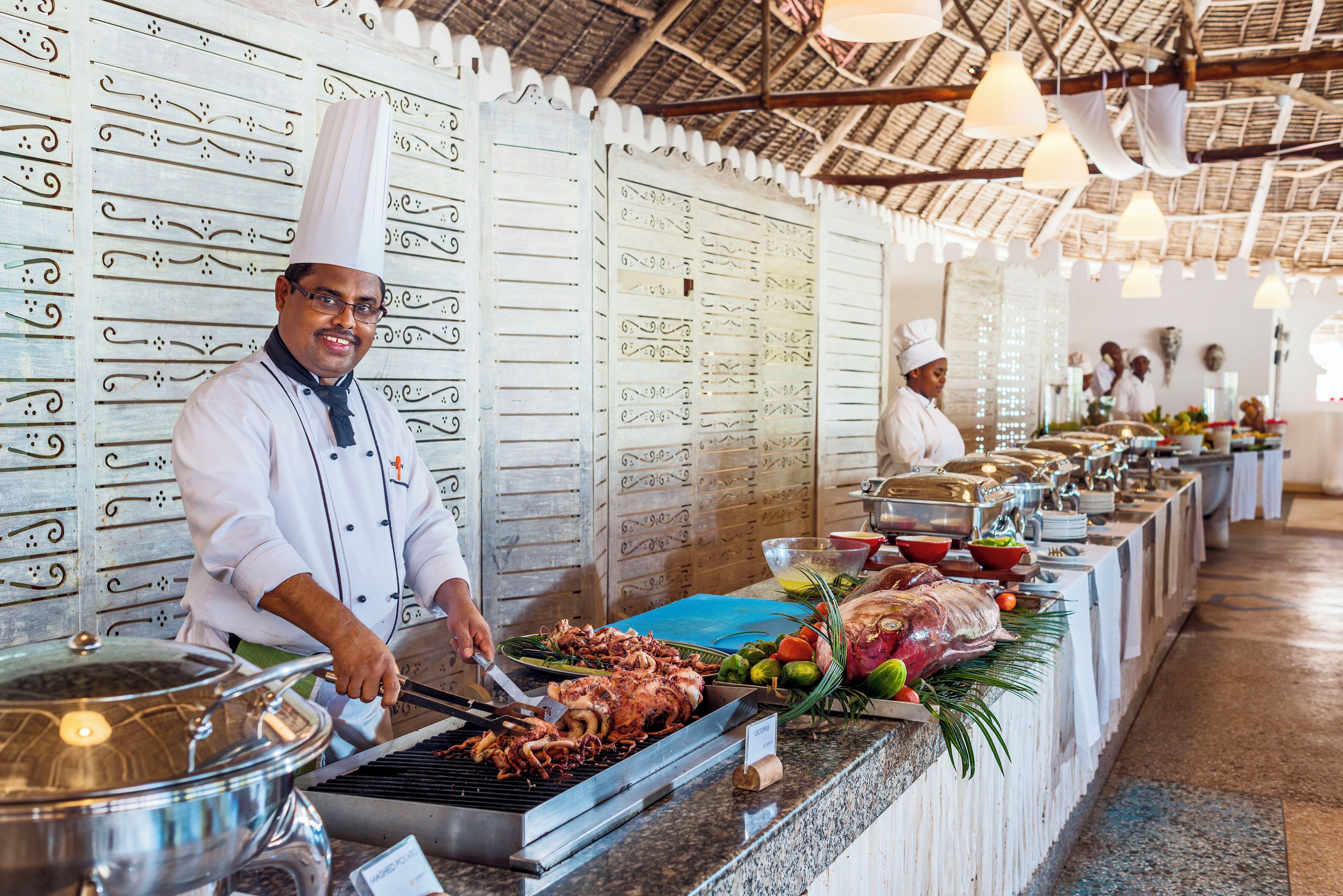 Disposição de buffet com um chefe preparando comida, variedade de pratos e uma parede decorativa com padrões.