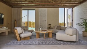 Uma sala de estar moderna com teto de madeira, janelas amplas, um sofá, poltronas, uma mesa de centro e um grande painel vertical com texto.
