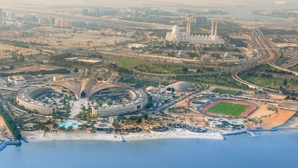 Vista aérea de uma cidade costeira com um estádio grande, uma mesquita e um moderno complexo hoteleiro com design circular e uma praia.