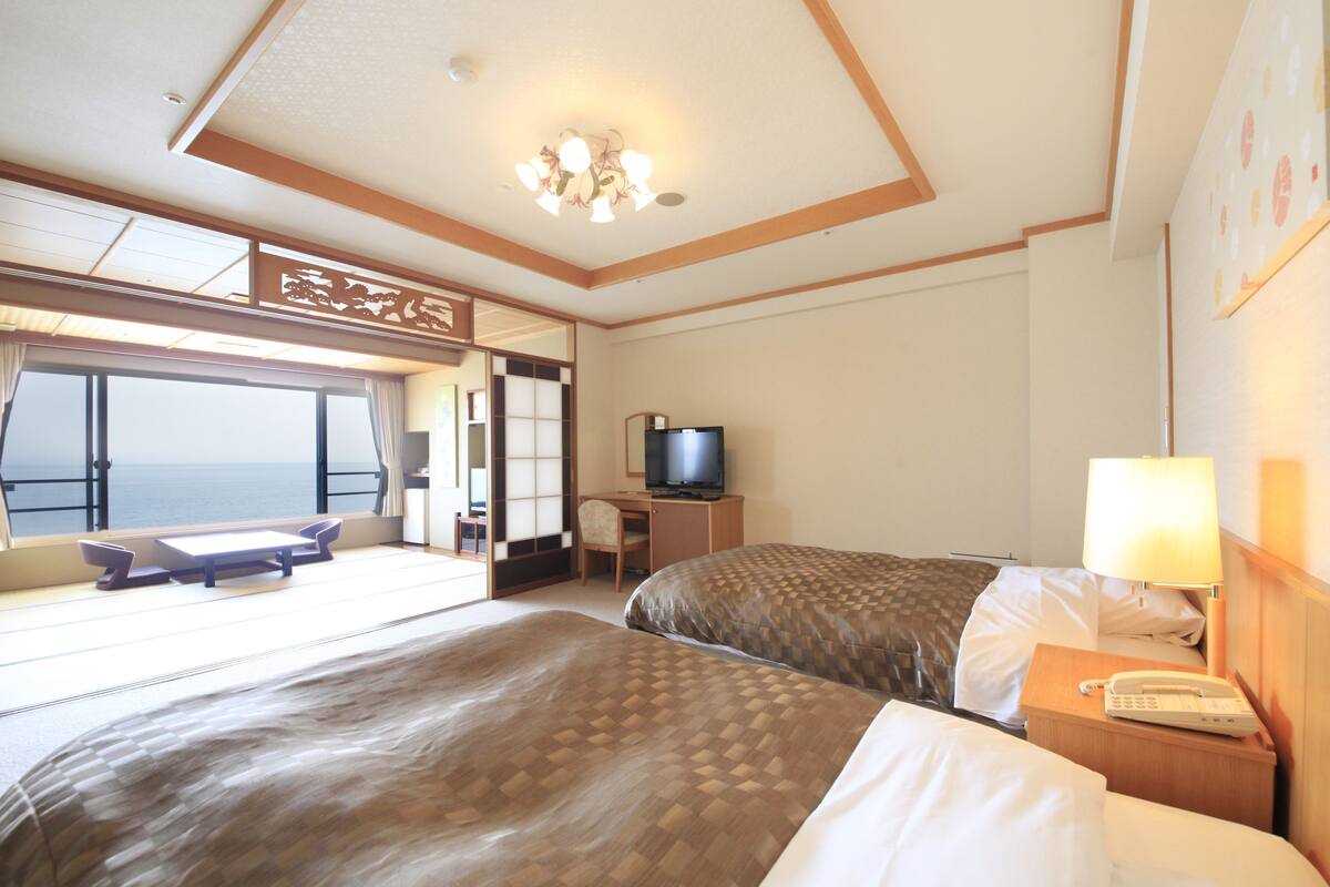 Quarto de hotel com duas camas, uma escrivaninha com televisão, uma luminária e vista para o mar.