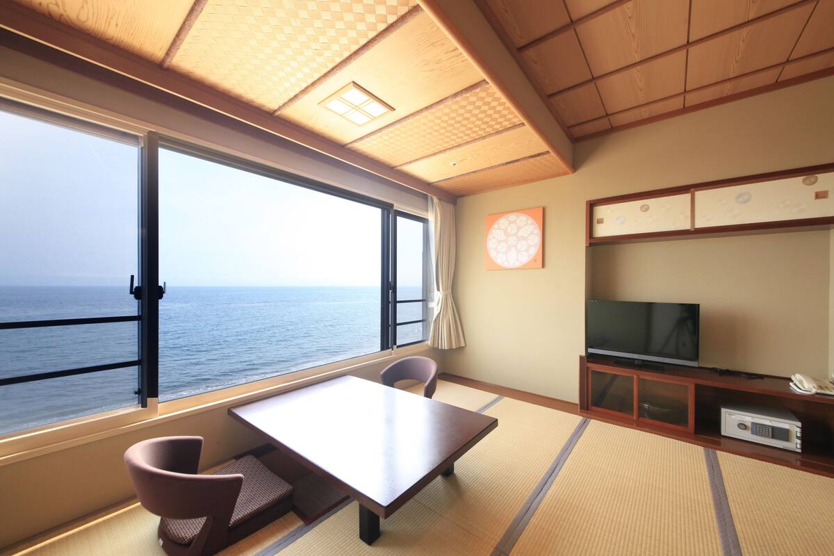 Quarto com uma grande janela com vista para o mar, uma televisão e uma pequena mesa com cadeiras.