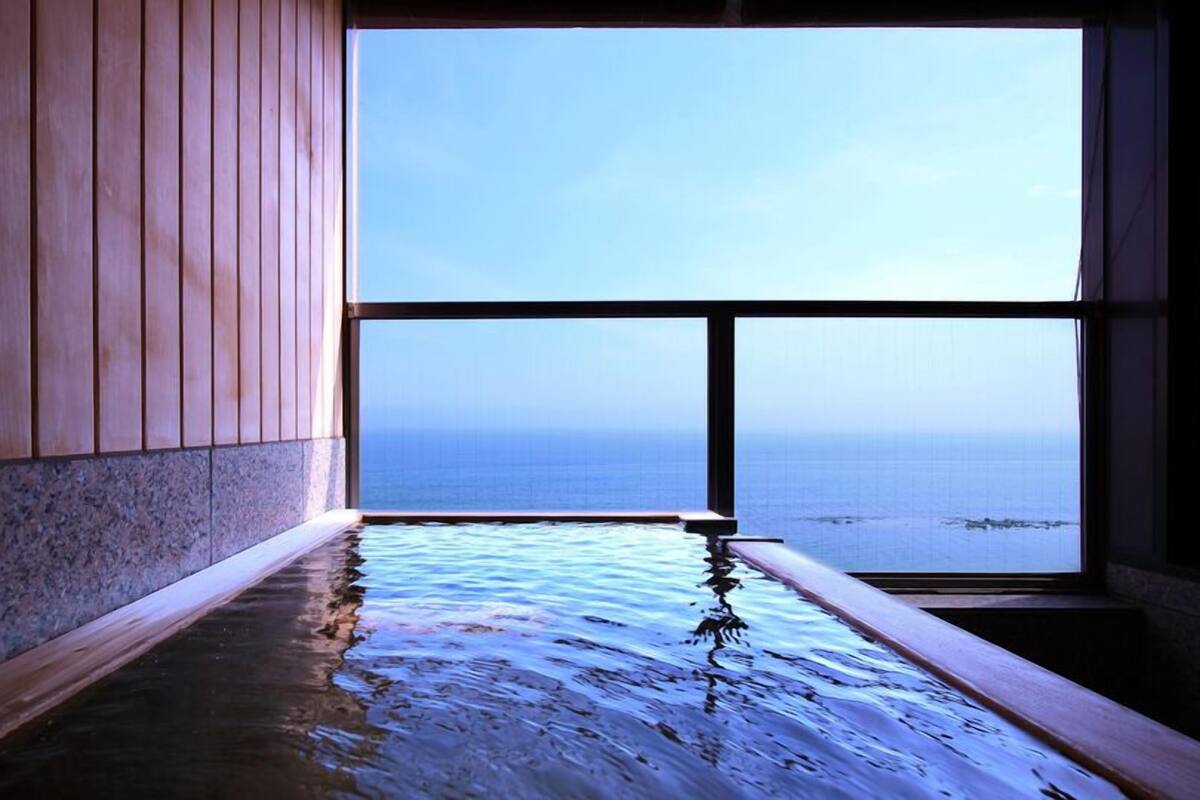 Quarto com uma grande janela, piso de madeira e vista para o mar.