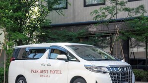 Uma minivan branca com a inscrição 'President Hotel Hakata' estacionada em frente a um edifício.