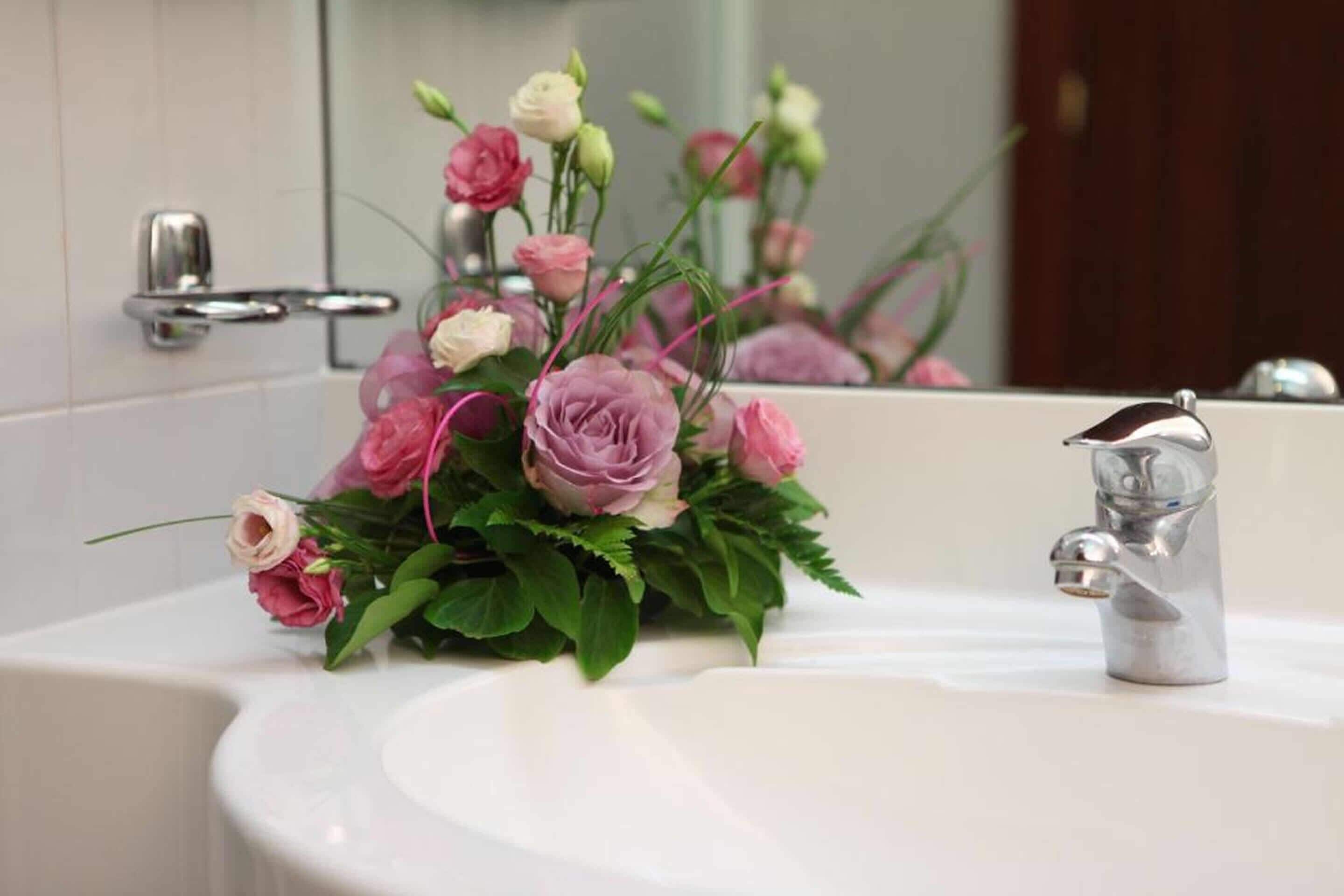 Um banheiro com pia branca, torneira prateada e um espelho refletindo um buquê de flores rosa e branco.