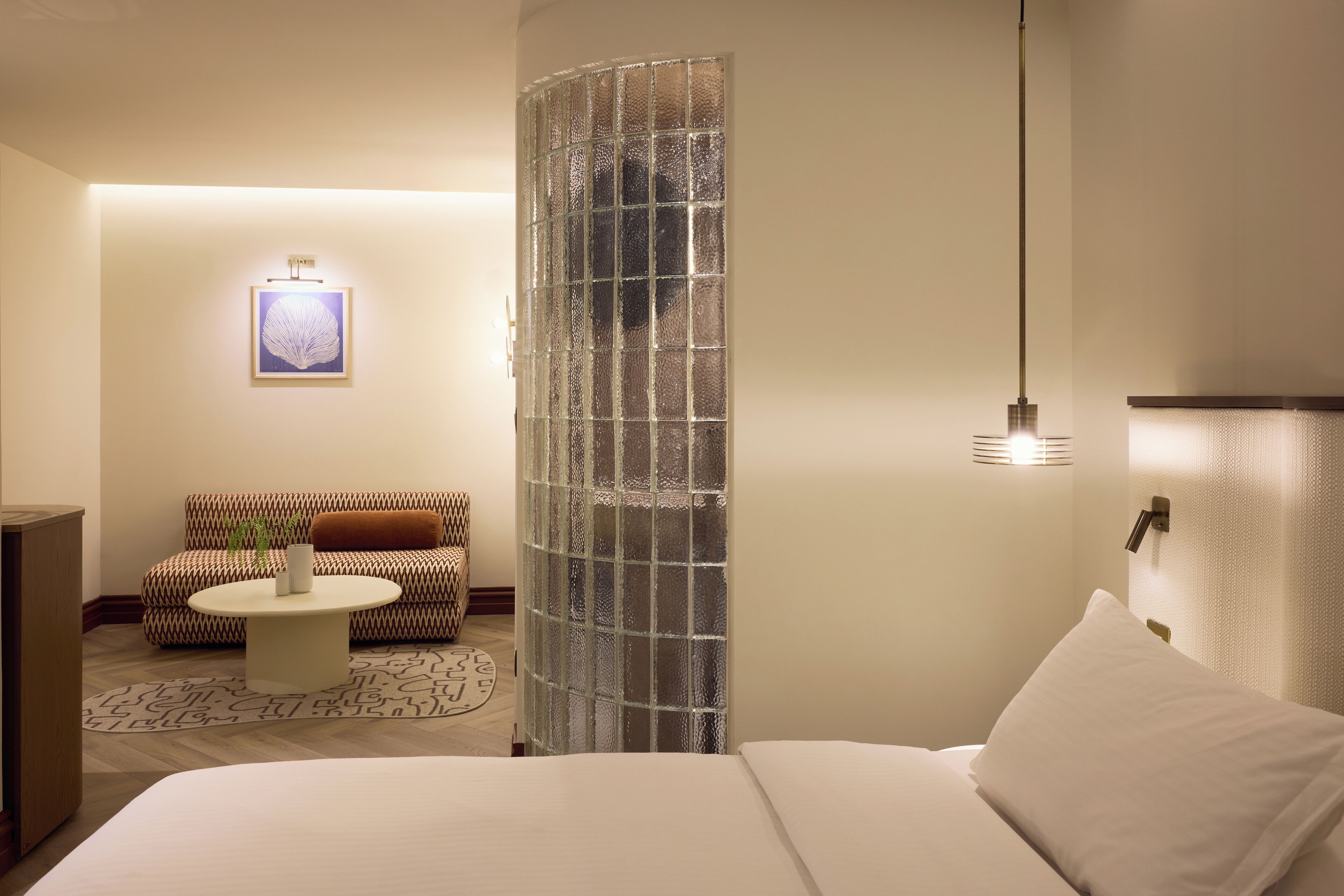 Quarto de hotel com cama, mesa de centro redonda, sofá com almofadas estampadas, quadro na parede e divisória de vidro decorativa.