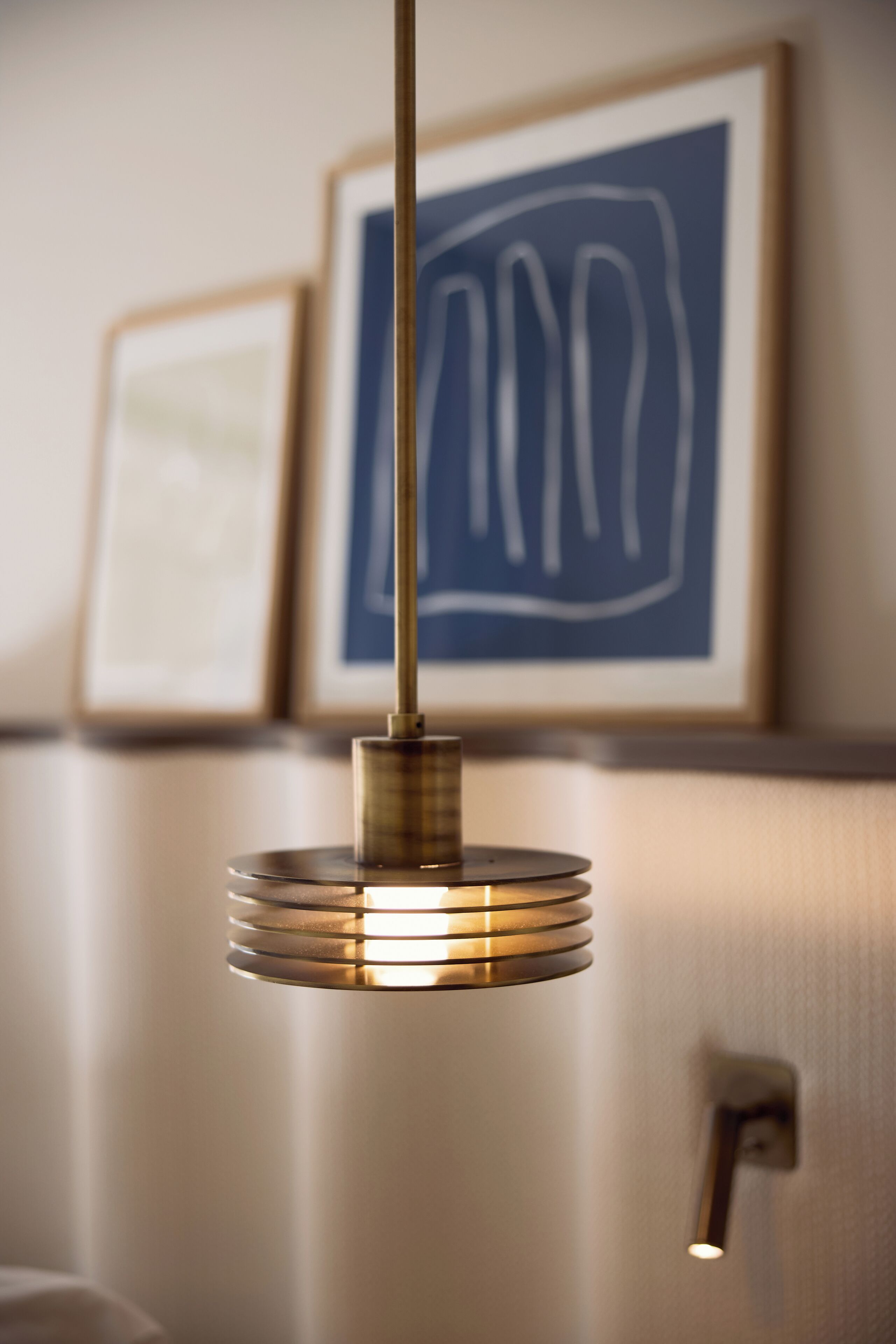 Uma luminária pendente com acabamento metálico, suspensa no teto de um quarto com obras de arte emolduradas na parede.