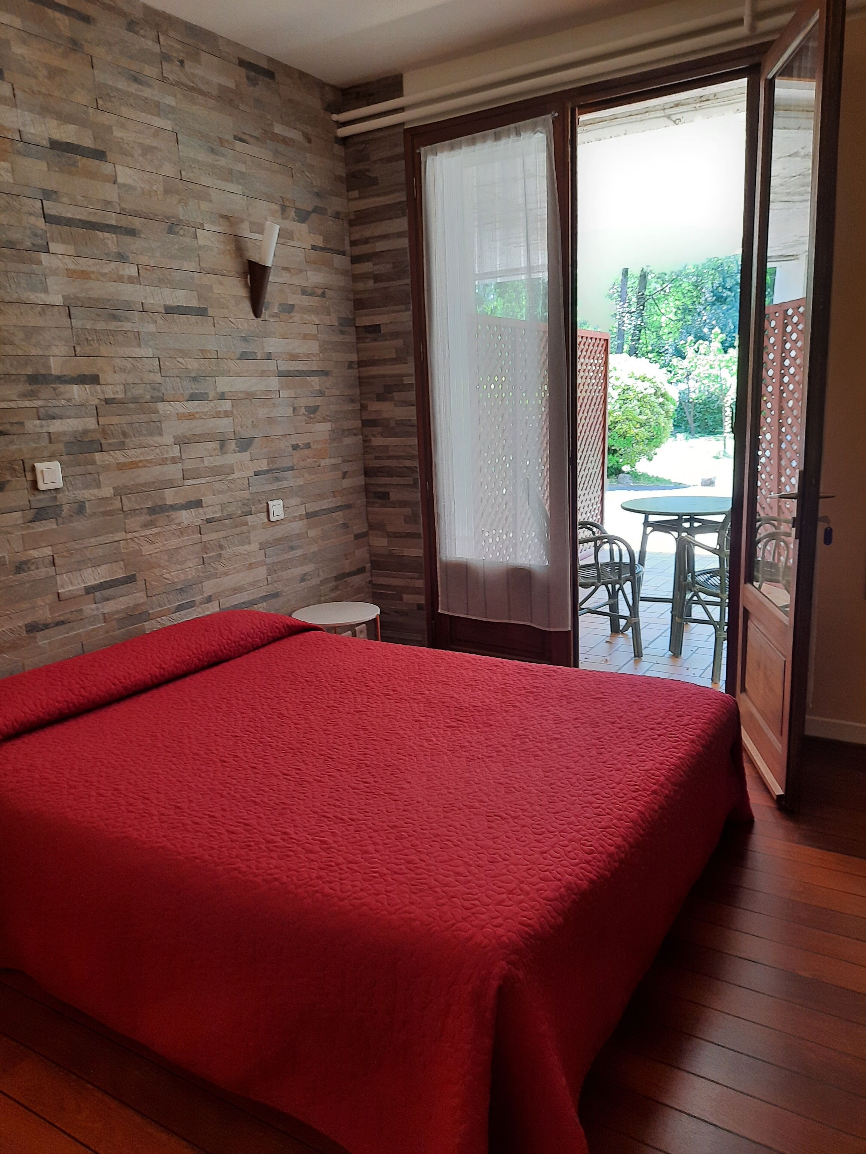 Quarto com colcha vermelha, parede de madeira e porta de vidro deslizante para um pátio externo com mesa e cadeiras.