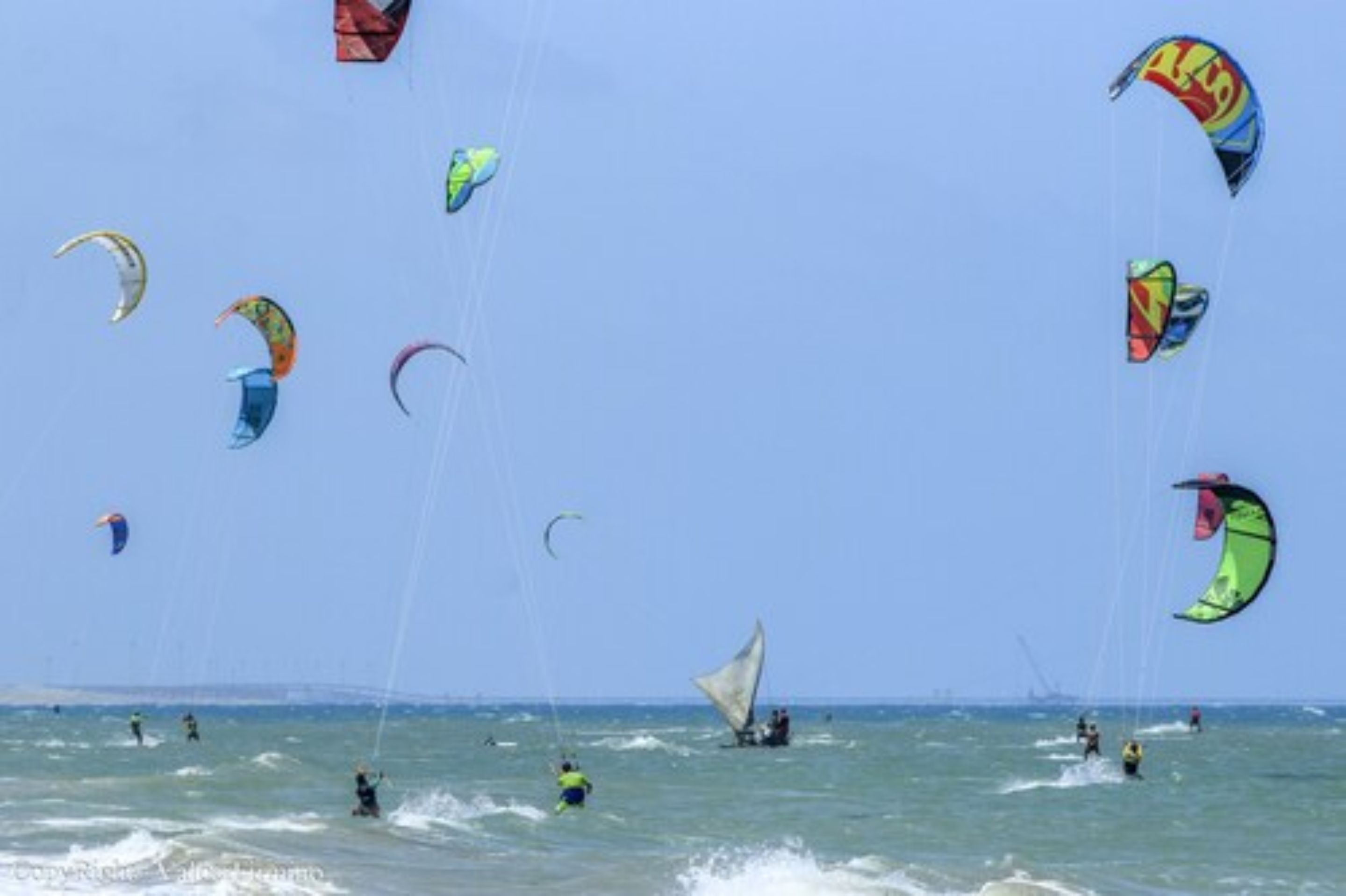 Uma praia com várias pessoas praticando windsurf e parasailing.