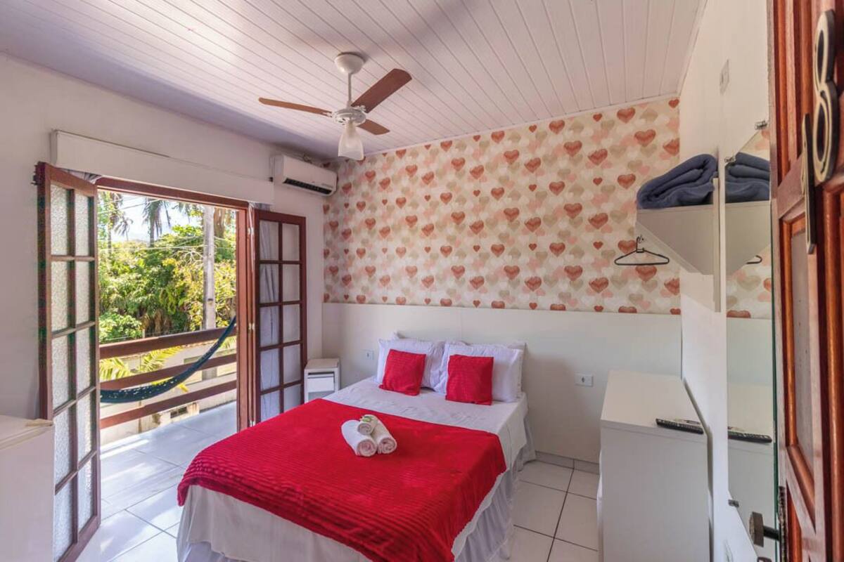 Quarto de hotel com cama, ventoinha no teto, janela com vista para o verde e parede com desenho de coração.