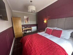 Quarto de hotel com cama grande, guarda-roupa de madeira, escrivaninha com cadeira, espelho e candelabro.