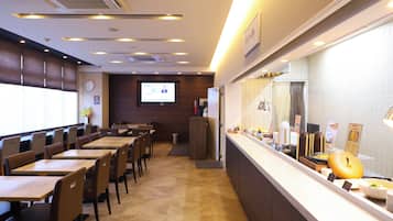 Um restaurante com mesas e cadeiras de madeira, um balcão com alimentos e uma tela de televisão ao fundo.