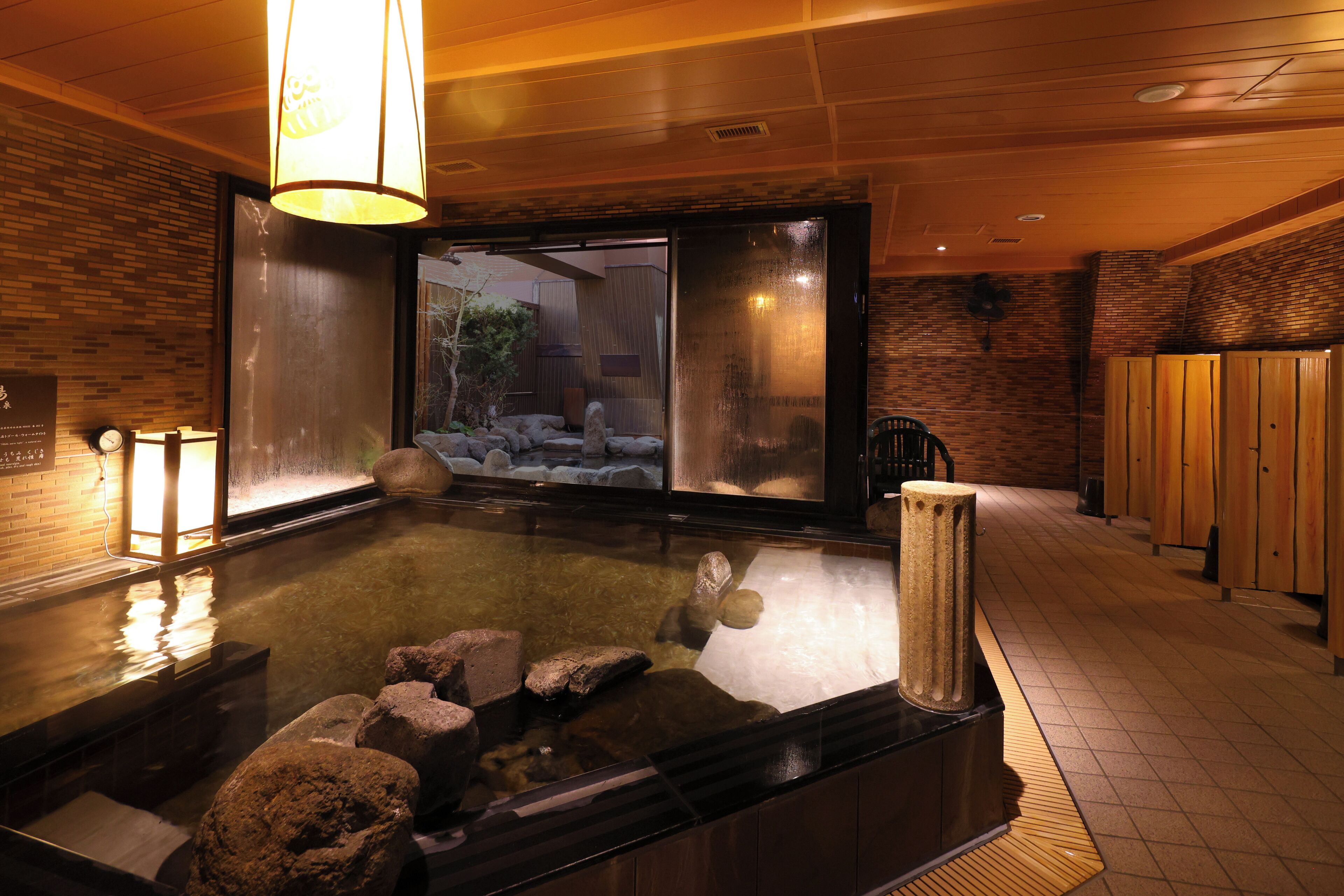 Uma onsen japonesa tradicional com interior em madeira, banho de águas termais e vista para um jardim através de uma divisória de vidro.