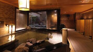 Uma onsen japonesa tradicional com interior em madeira, banho de águas termais e vista para um jardim através de uma divisória de vidro.