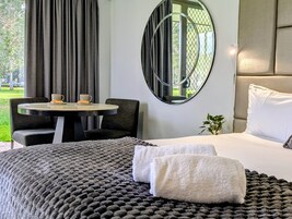 Quarto de hotel com cama, espelho redondo, mesa e duas cadeiras, e vista para o verde lá fora.