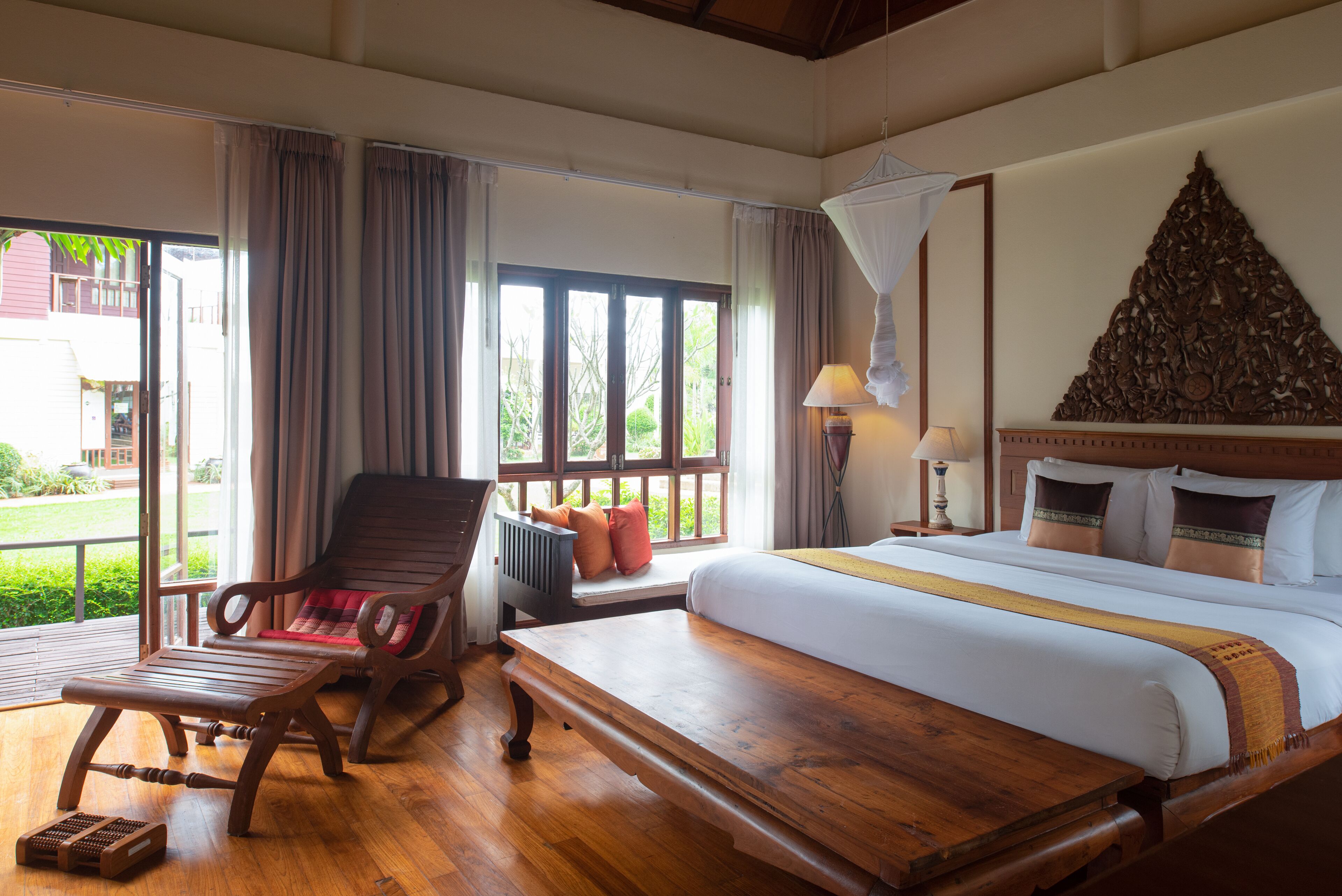 Quarto com uma cama grande, um banco de madeira, uma janela com cortinas e vista para o verde lá fora.