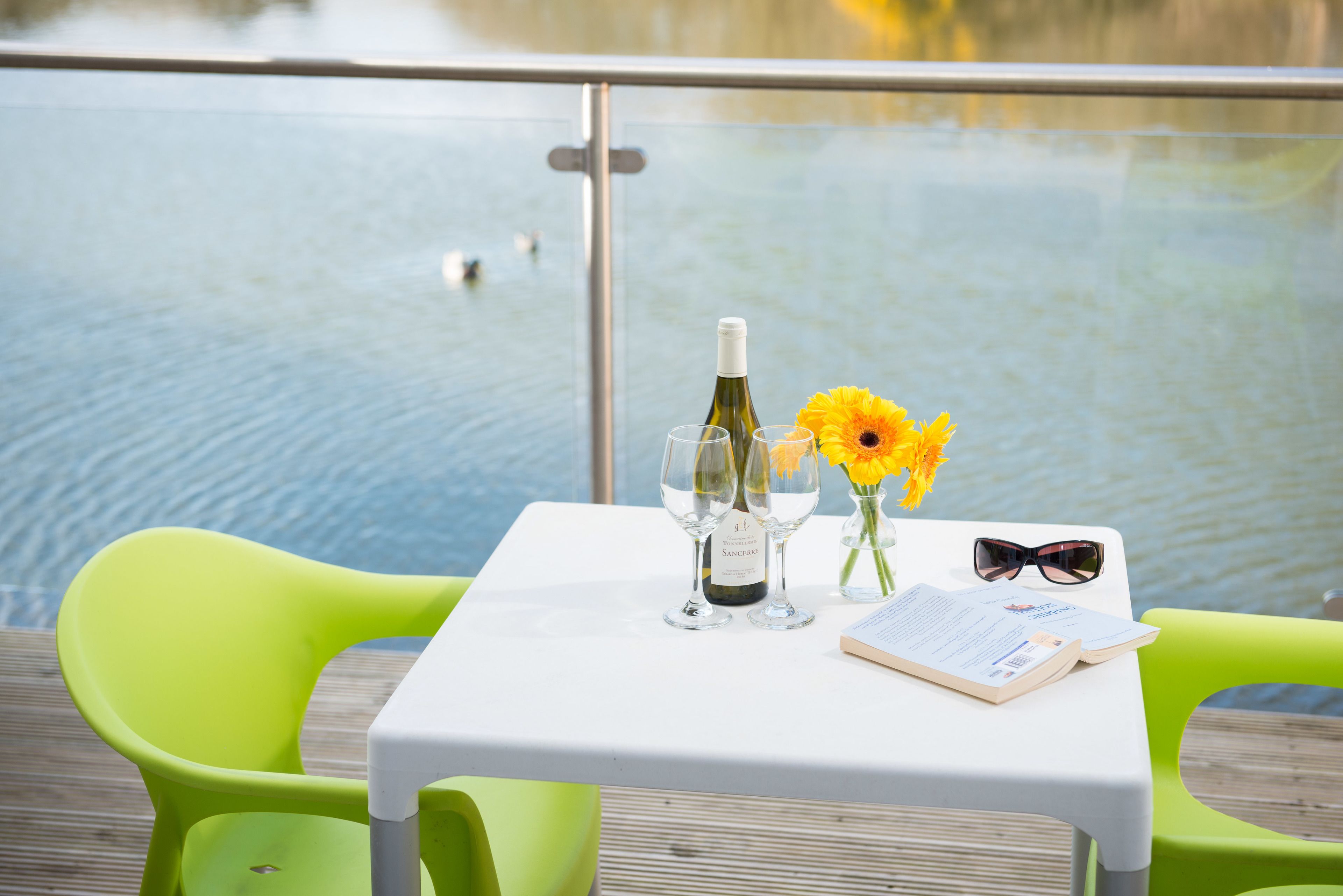 Uma mesa com uma garrafa de vinho, dois copos, um livro e óculos de sol, com vista para um lago calmo onde um cisne nada.