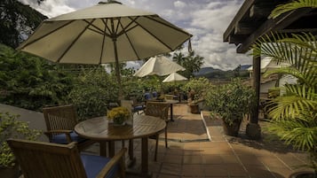 Um pátio externo com mesas e cadeiras de madeira, rodeado por plantas em vasos e guarda-sóis.