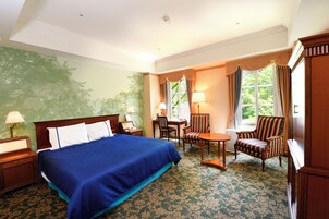 Quarto de hotel com uma cama grande, uma escrivaninha, duas cadeiras, um abajur e vista para o verde pela janela.
